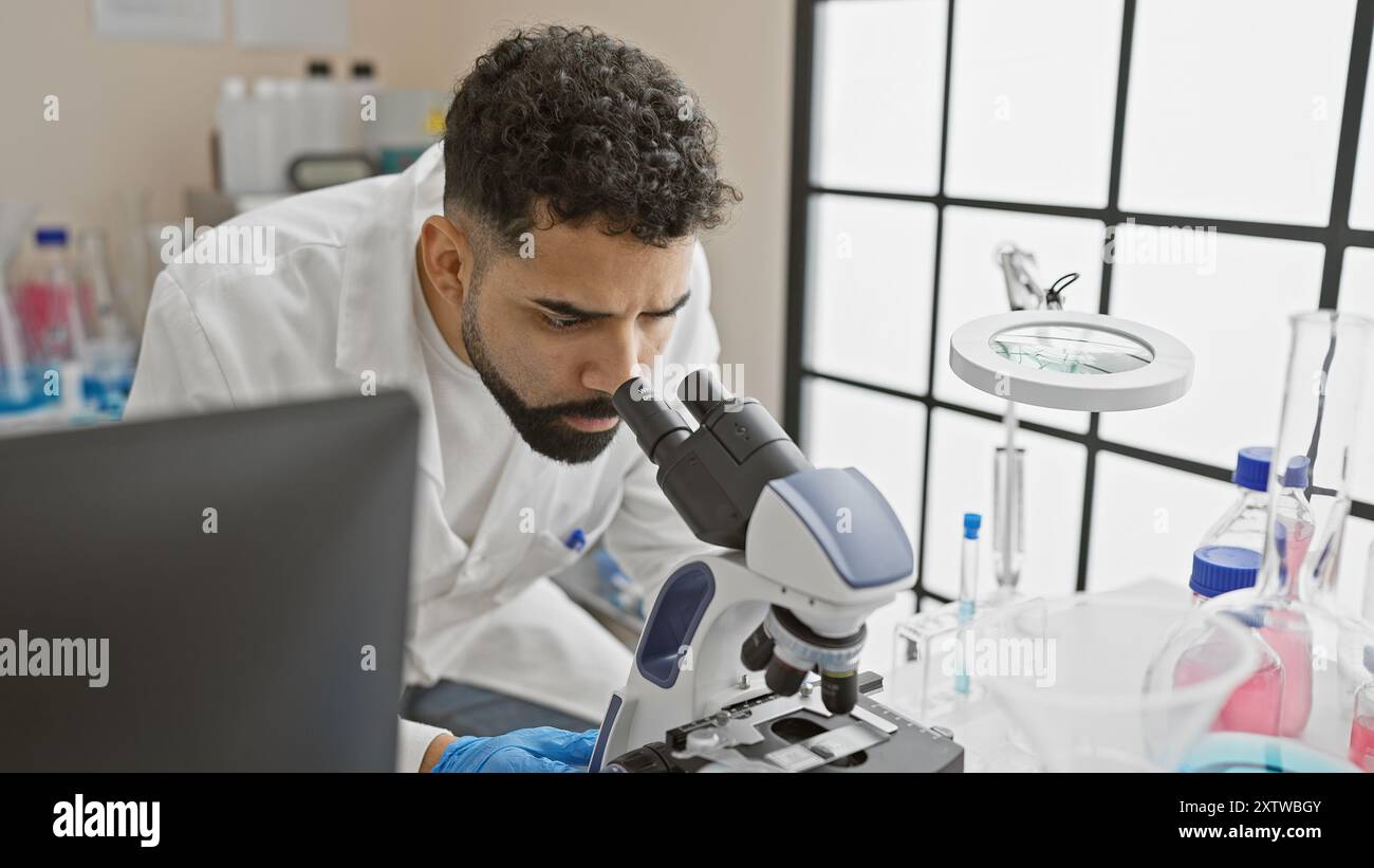 A focused hispanic man with a beard examines samples using a microscope ...