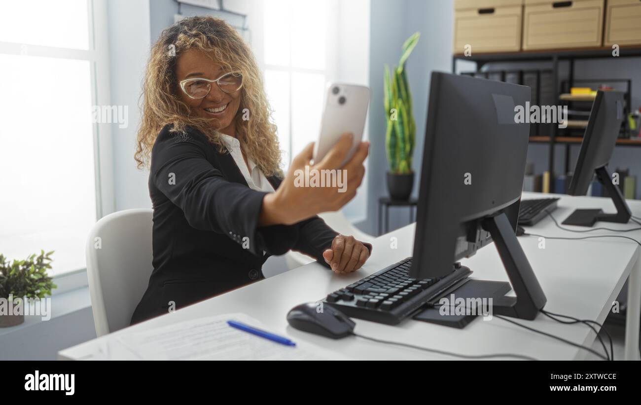 Woman taking selfie indoor at office desk with computer and plant in ...