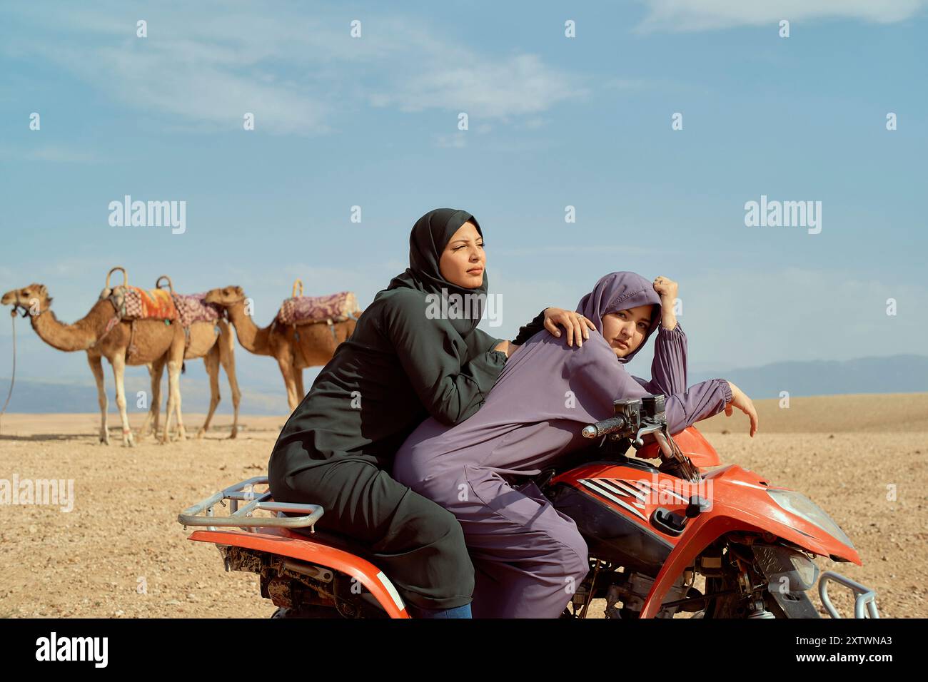 Two women in traditional clothing resting on an ATV in a desert landscape with a caravan of ...