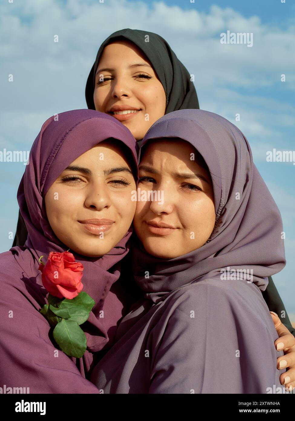 Three smiling women wearing hijabs embrace against a blue sky, one ...