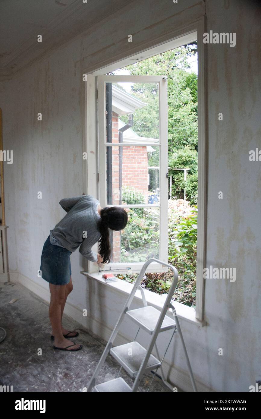 A woman in casual attire is renovating a house, scraping paint from a ...