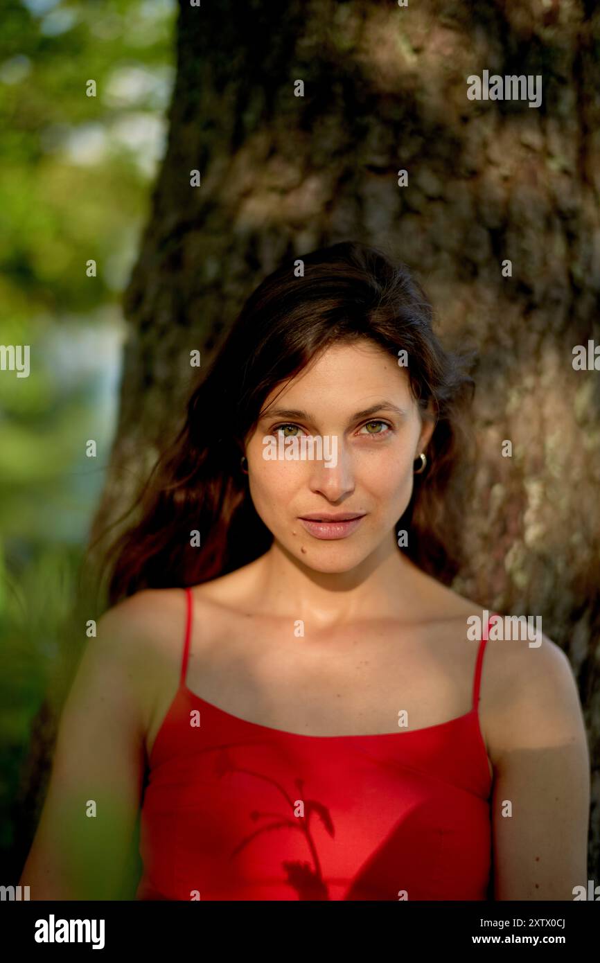 Woman in red dress standing in front of a tree with sunlight filtering through the leaves Stock ...
