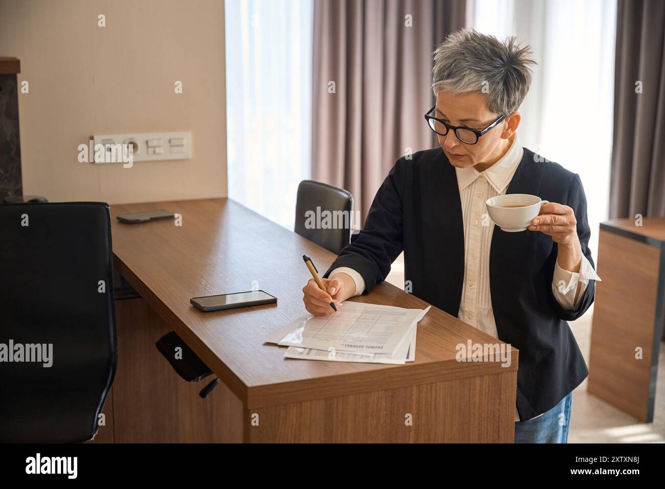 Senior lady filling out the form, holding cup of tea in hotel room ...