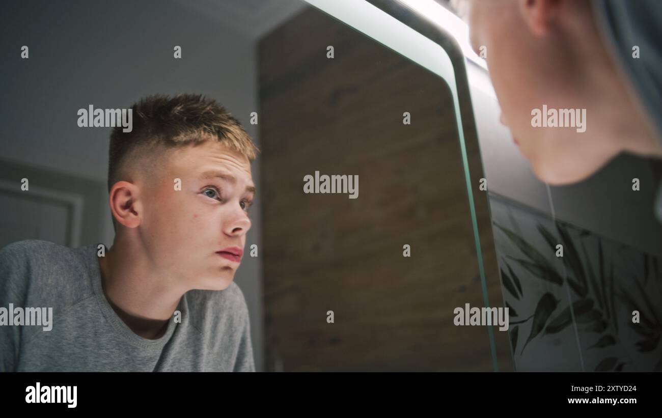 Teenager stands near wash basin and touches his face in the morning ...