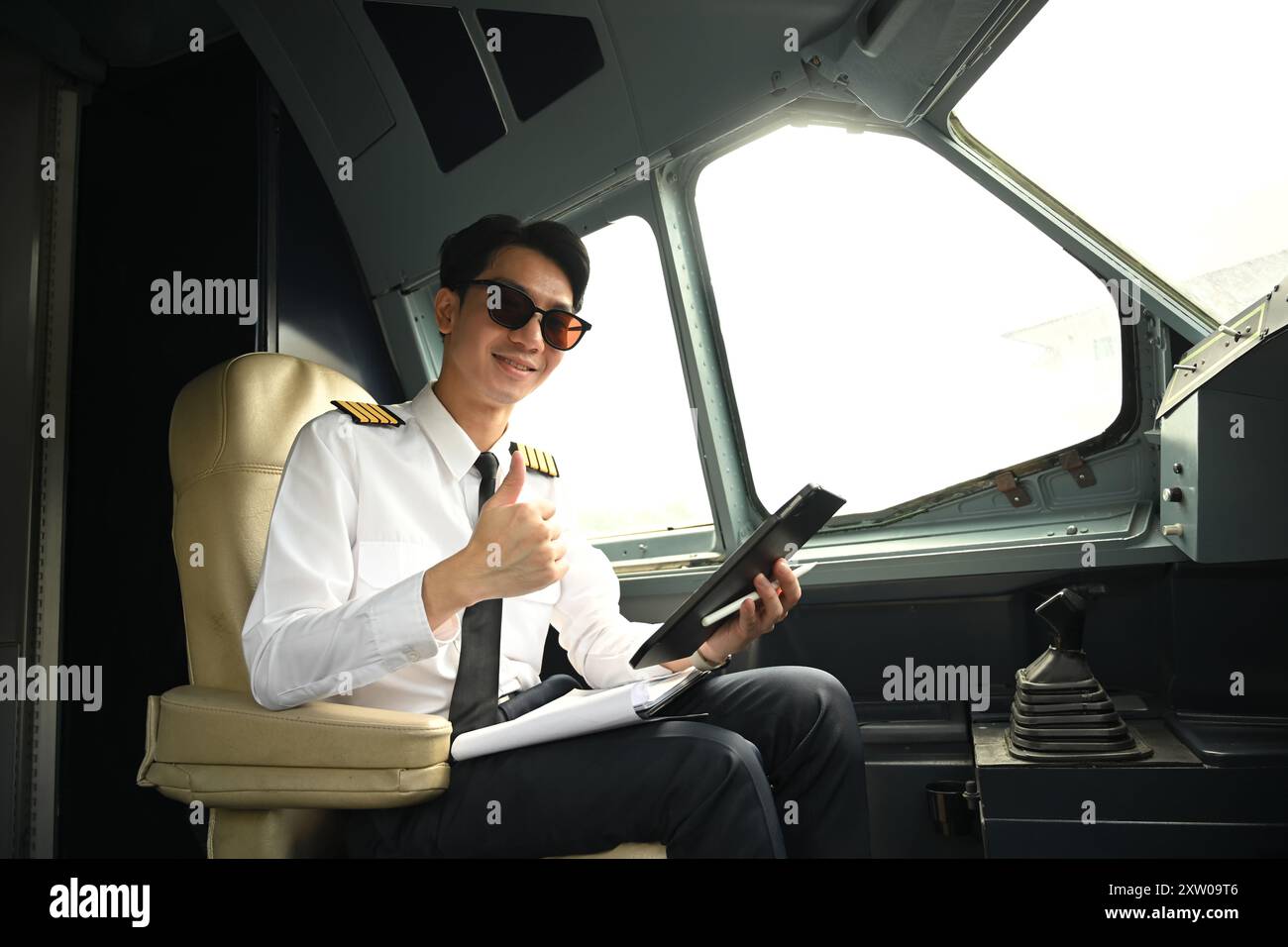 Confident professional airline pilot showing thumb up while sitting in the cockpit of an ...