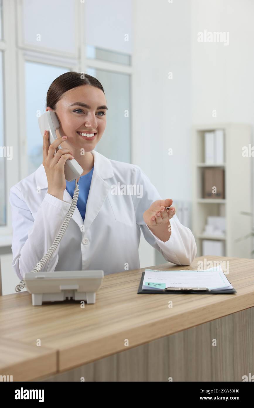 Professional receptionist talking on phone at wooden desk in hospital ...