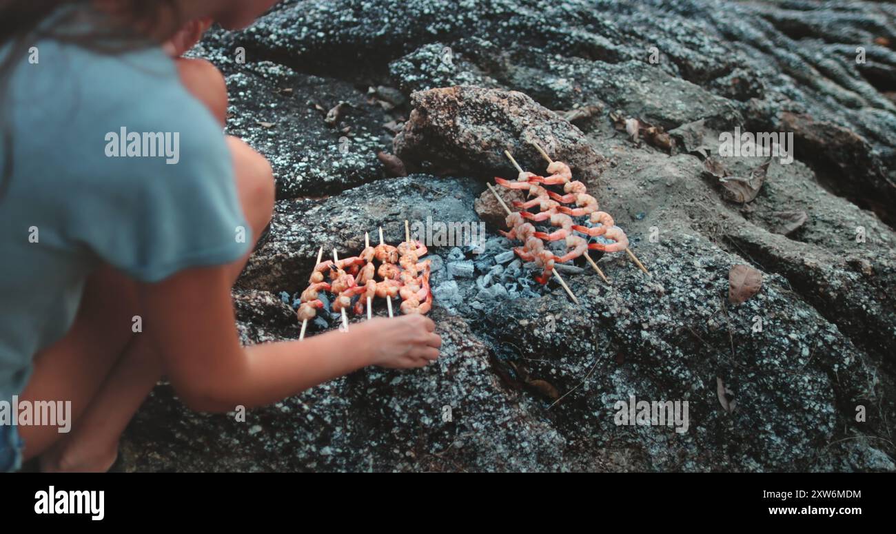 Woman is grilling shrimp skewers on a makeshift grill made of rocks in ...