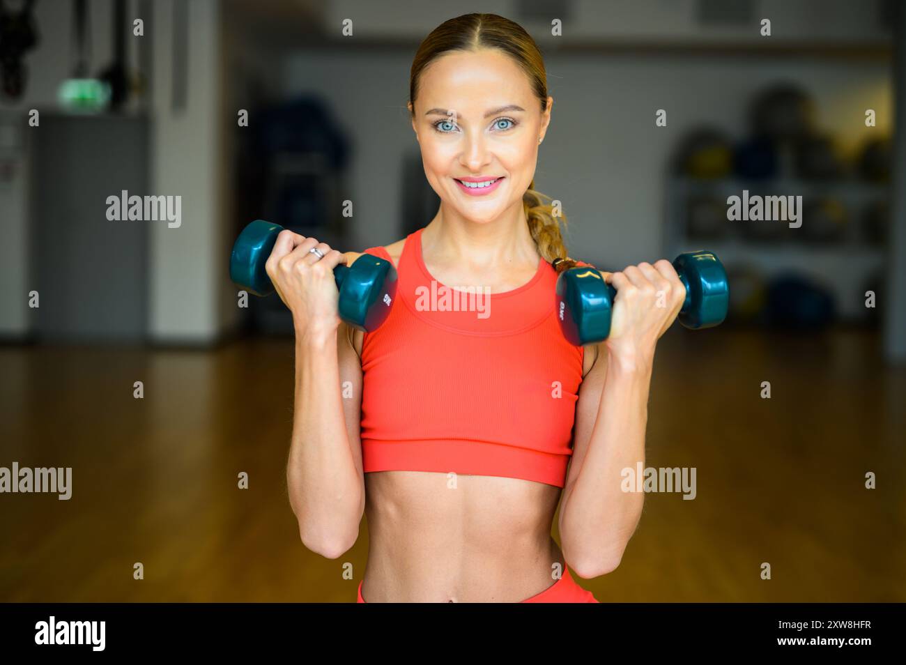 Smiling athletic woman in red workout gear lifting weights indoors ...
