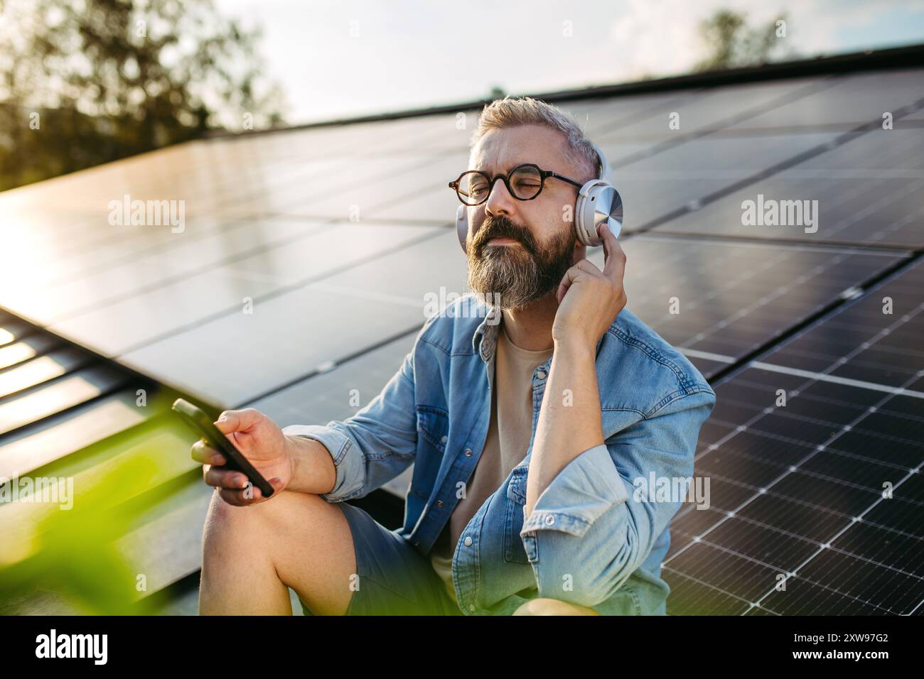 Man sitting on roof full of solar panels, headphones on head and closed ...