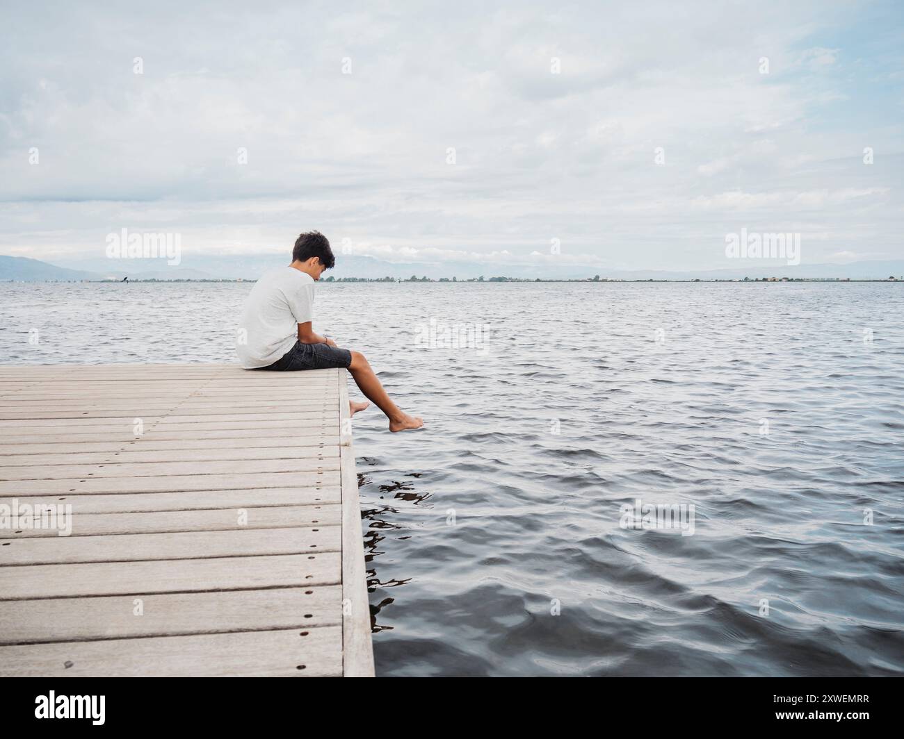 Young boy sitting alone on the edge of a wooden pier, dipping their ...