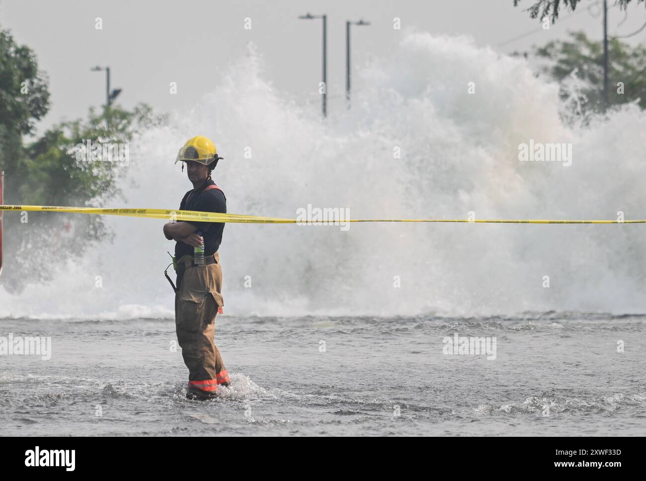 A firefighter stands on a flooded street next to a geyser caused by a ...