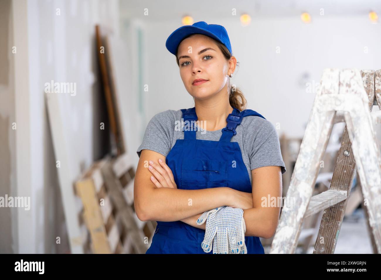 Portrait of positive builder woman in blue overalls next to stepladder ...