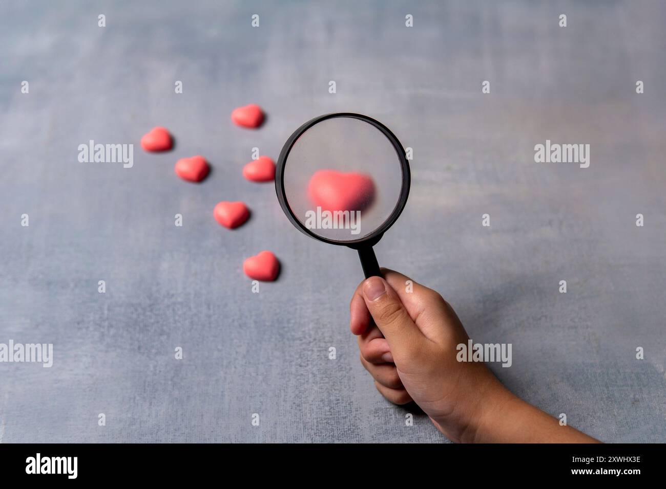A hand holding a magnifying glass focuses on a single pink heart among ...