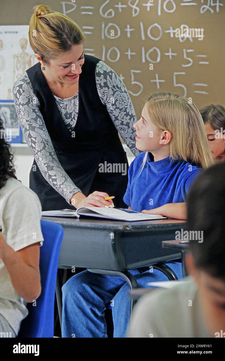 A teacher assists a student at a desk while other children work in a ...
