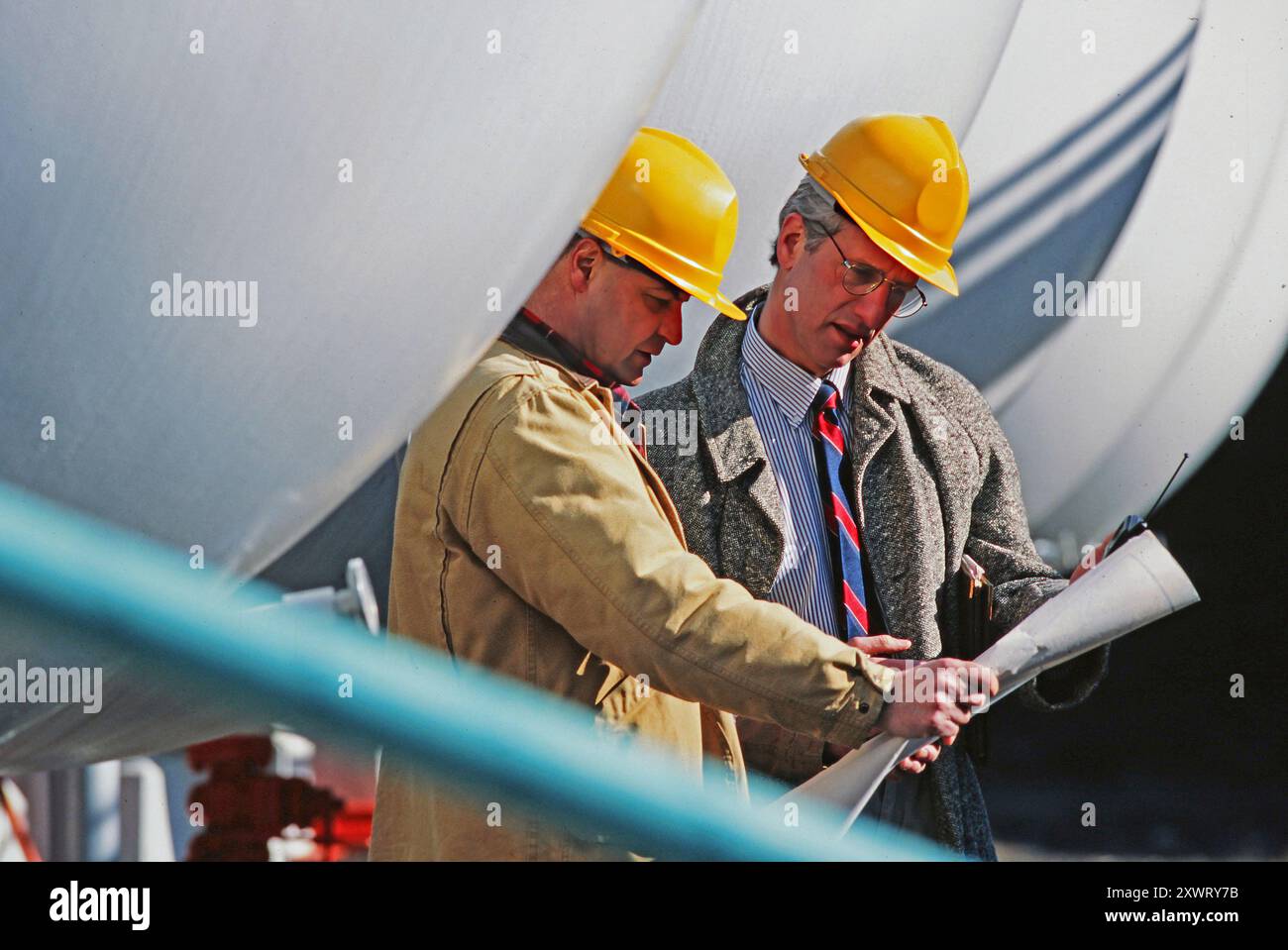 Engineers analyze blueprints while standing near large industrial ...