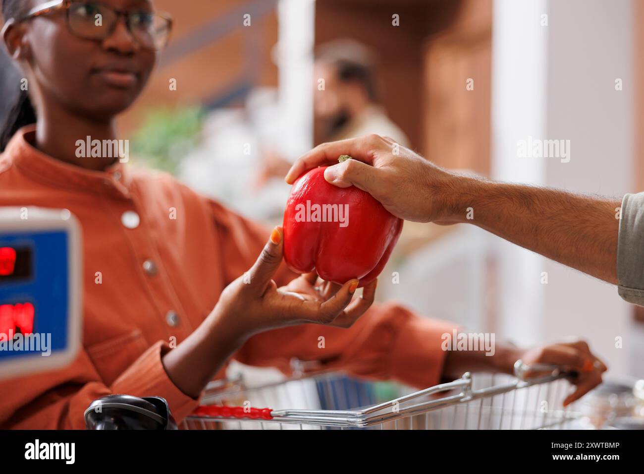 A black woman hands a fresh red bell pepper to a shopkeeper to weigh ...