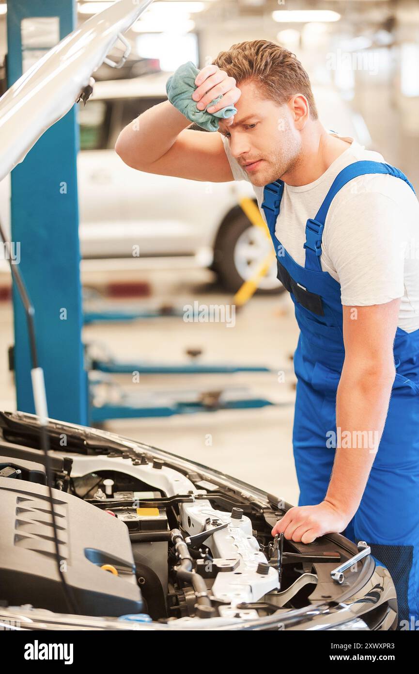 Tired mechanic. Tired young man in uniform examining car and wiping his ...