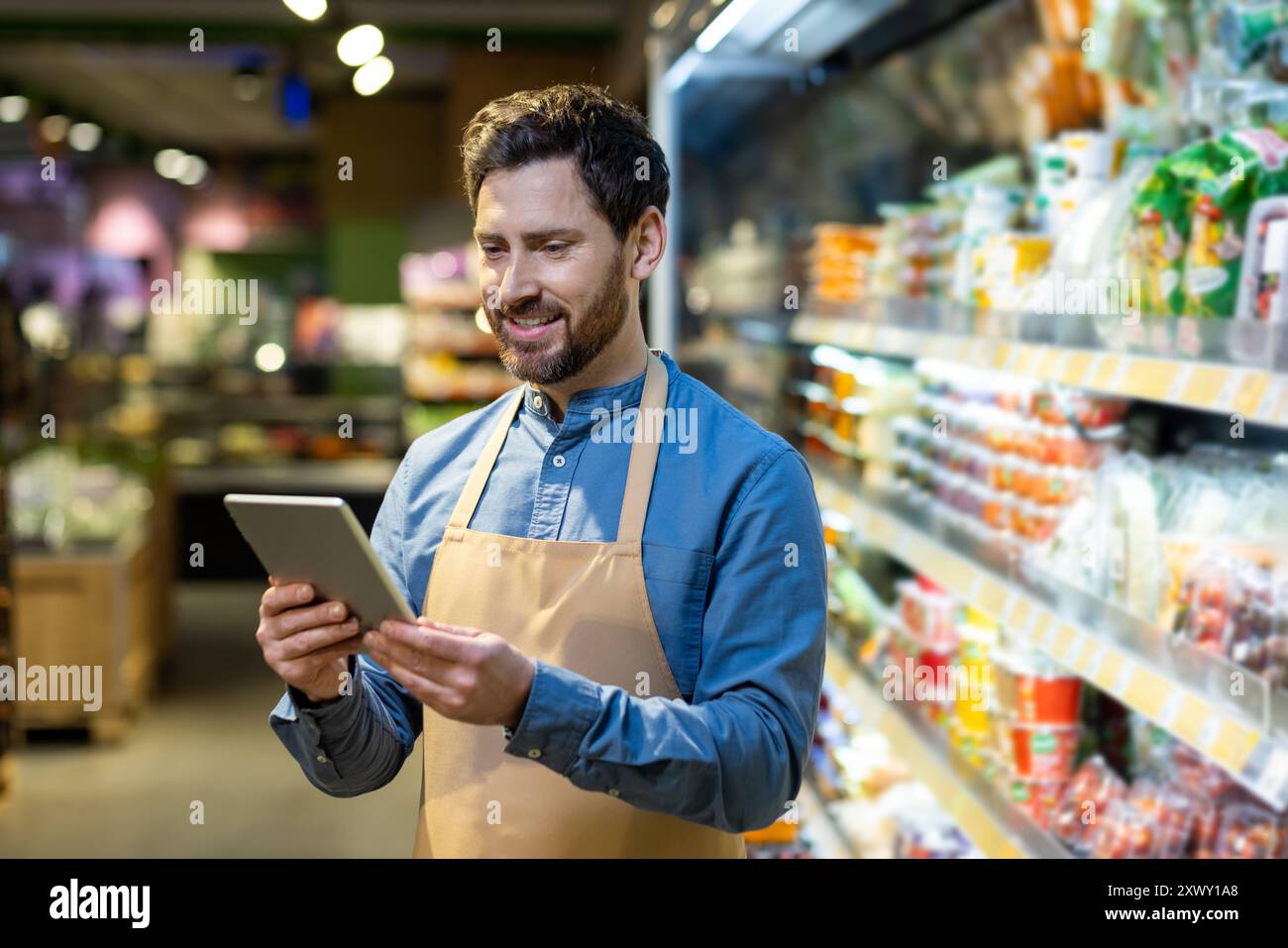 Grocery store employee wearing apron stands in supermarket aisle using ...