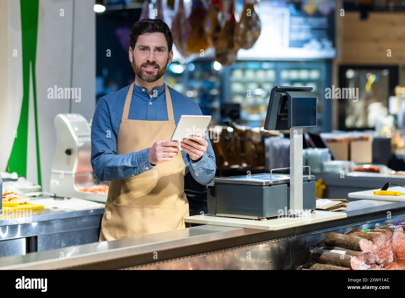 Professional shopkeeper in apron using tablet at delicatessen counter ...