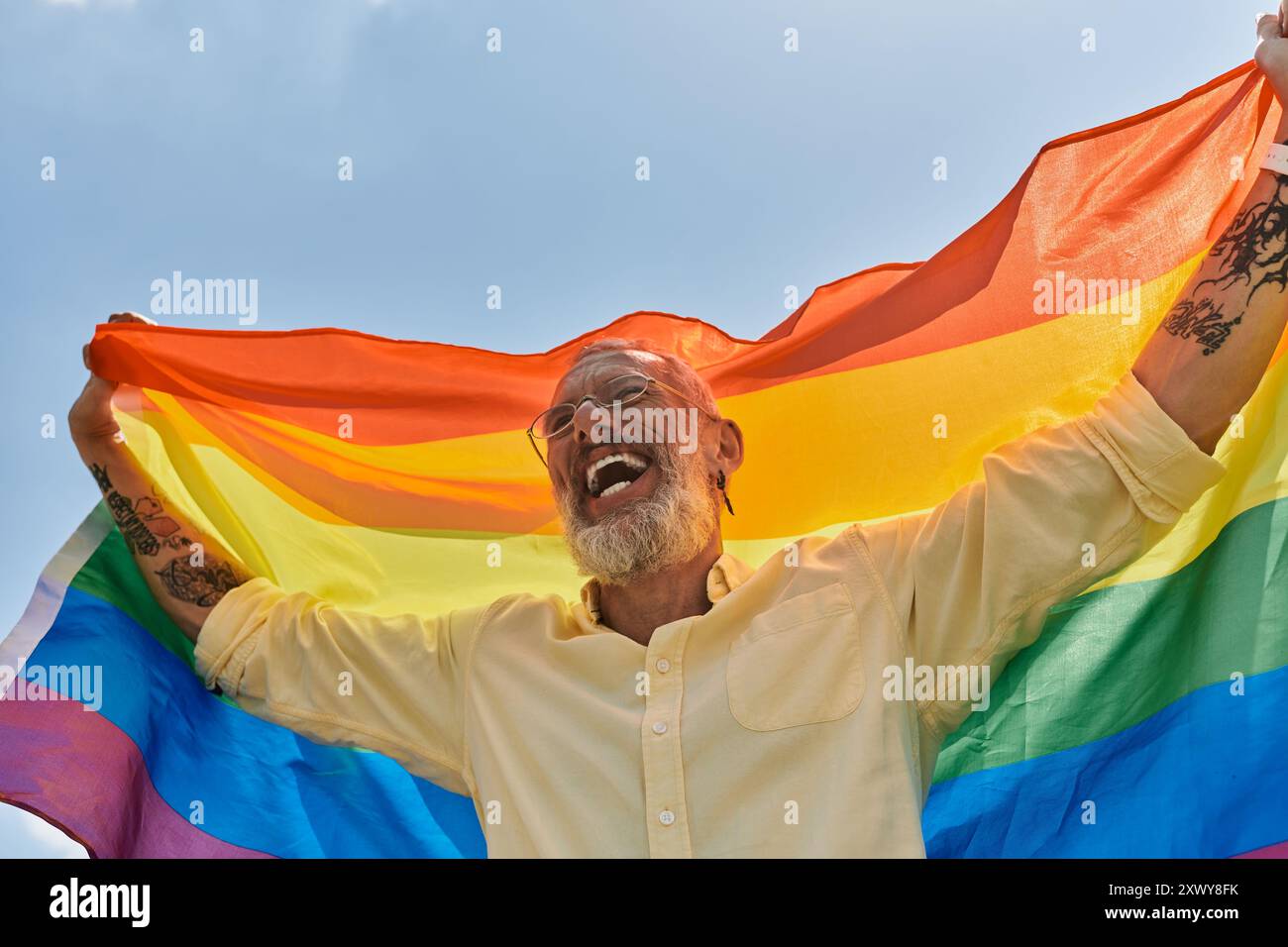 A middle-aged man with a beard holds a rainbow flag and laughs, beaming ...