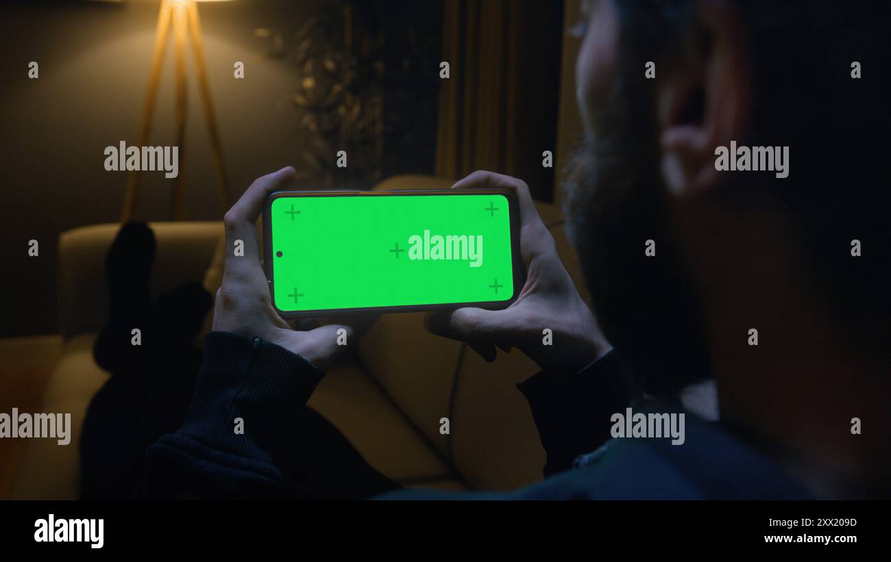 Man lying on couch using smartphone horizontally with chroma key green screen. Male watching ...