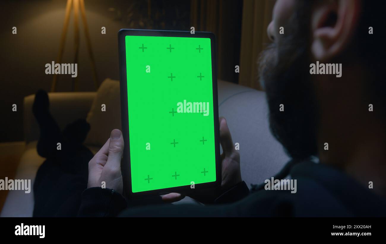 Back view of man lying on couch using tablet with chroma key green screen, scrolling through ...