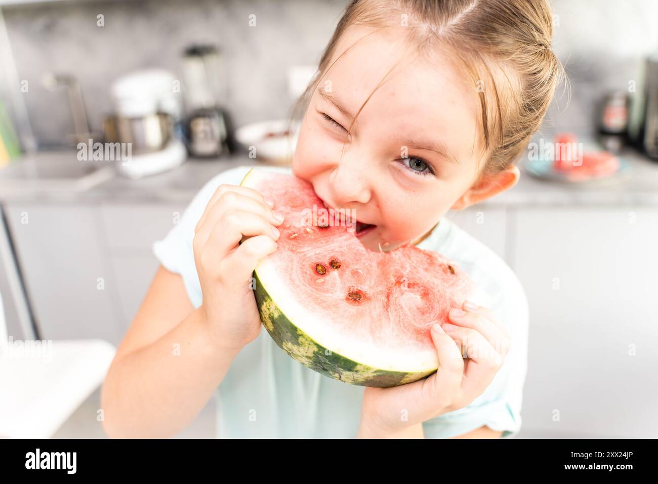 Little girl biting a piece of watermelon while standing in the kitchen ...
