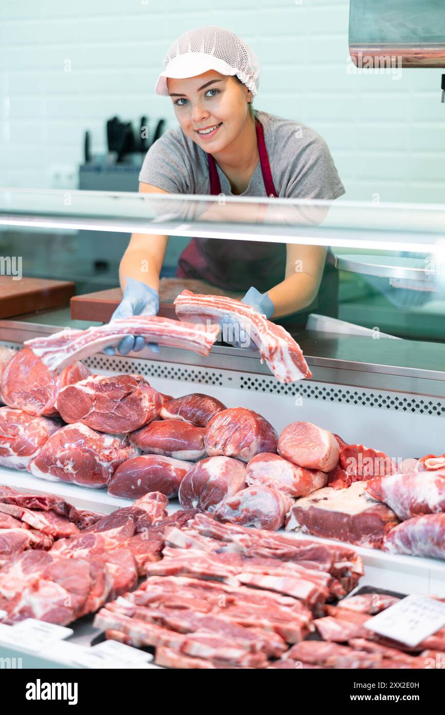 Young butcher shop saleswoman laying out raw beef ribs in display case ...