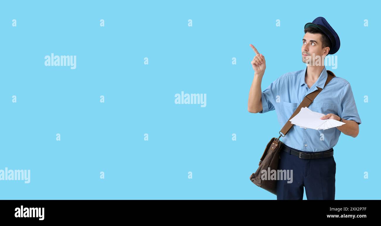Handsome young postman pointing at something on light blue background ...