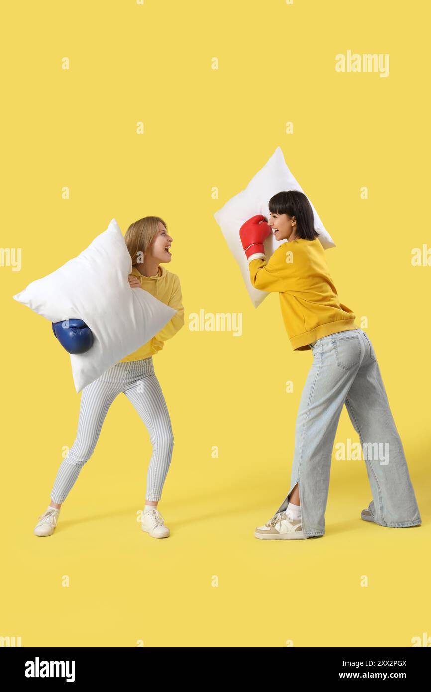 Young women in boxing gloves fighting pillows on yellow background ...