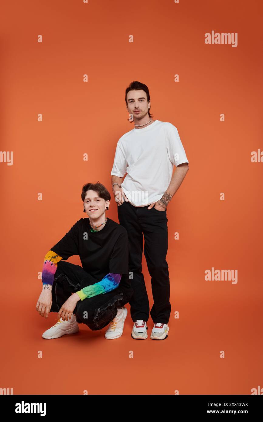 Two young queer friends pose in stylish attire against a vibrant orange ...