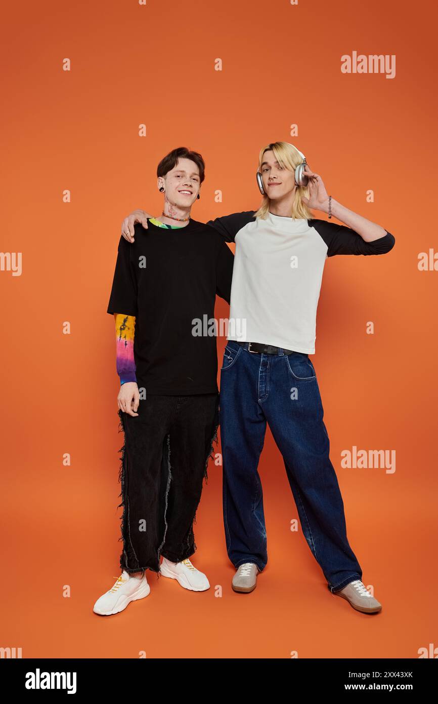Two young queer friends pose in stylish attire against a vibrant orange ...