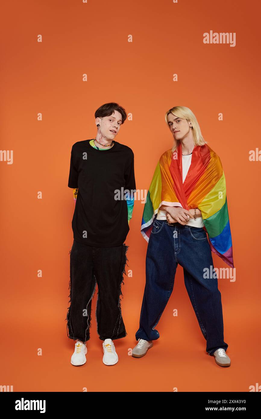 Two young friends, standing with rainbow flag, pose against an orange ...