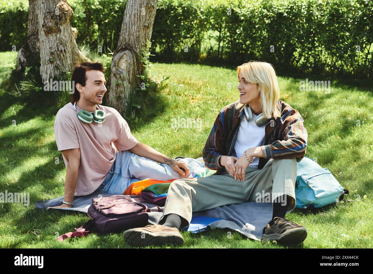 Two young queer friends sit on a blanket in a grassy park, laughing and ...