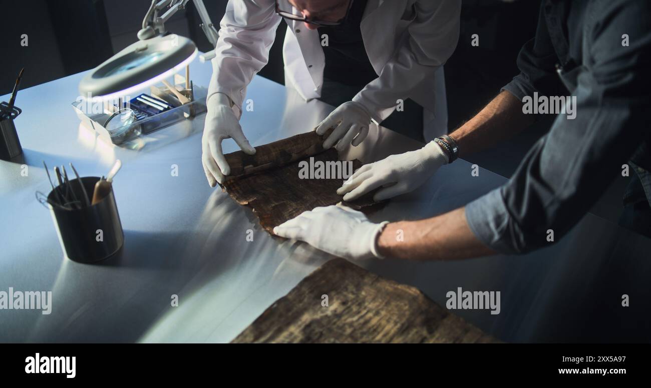 Two archaeologists in gloves carefully roll out ancient Egyptian papyrus on the table in ...