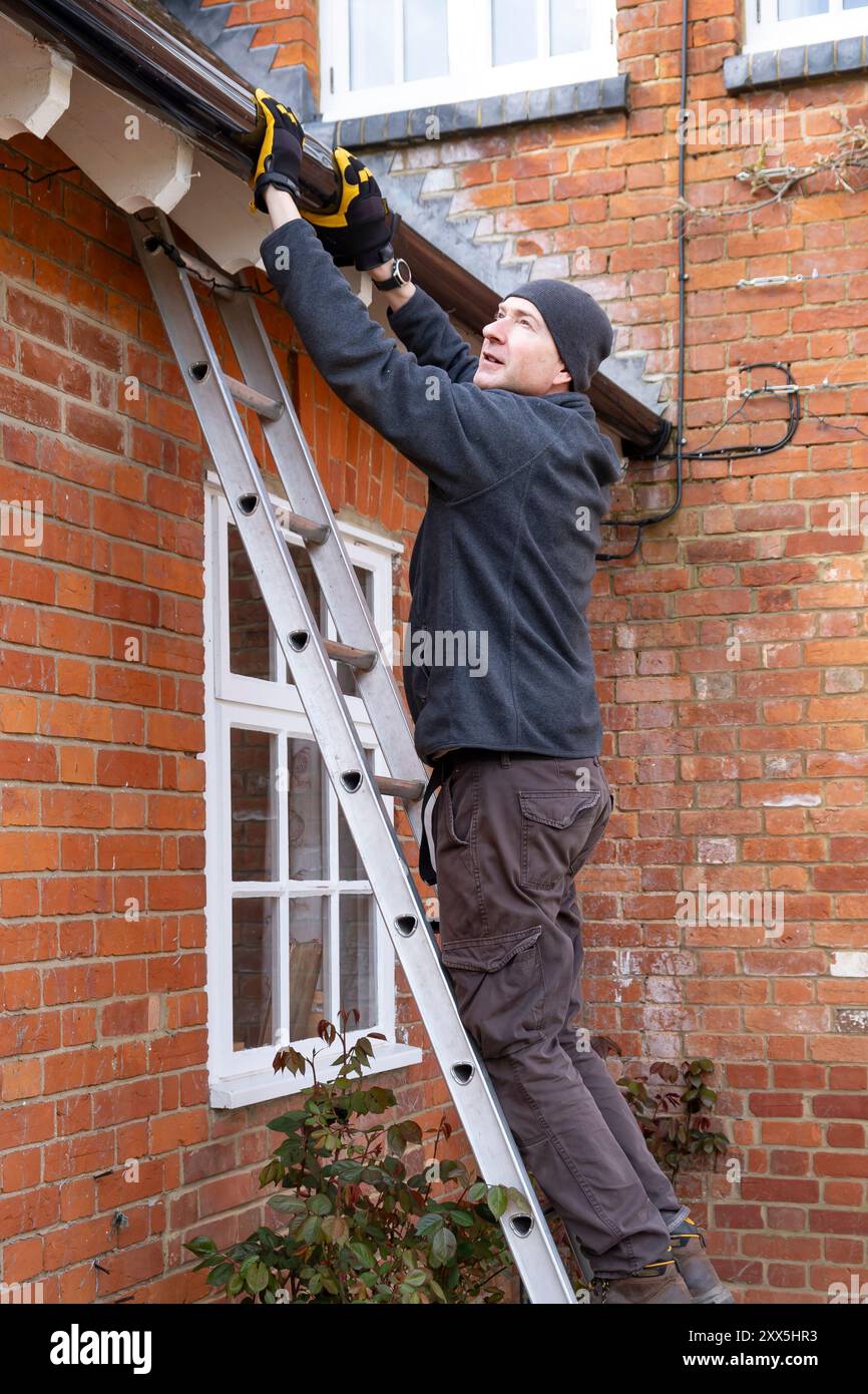 Man on a ladder fixing fitting a rain gutter on the eaves of a UK house ...