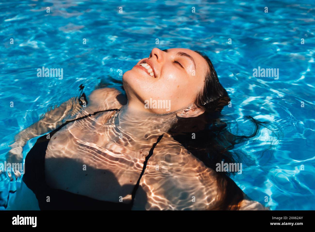 A young woman with long hair floats on her back in a pool, eyes closed, immersed in the ...