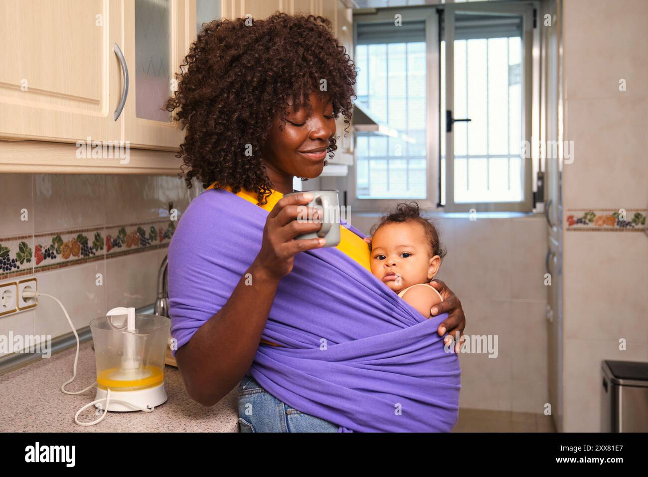 Mother is taking a break in her kitchen while holding her baby close to ...