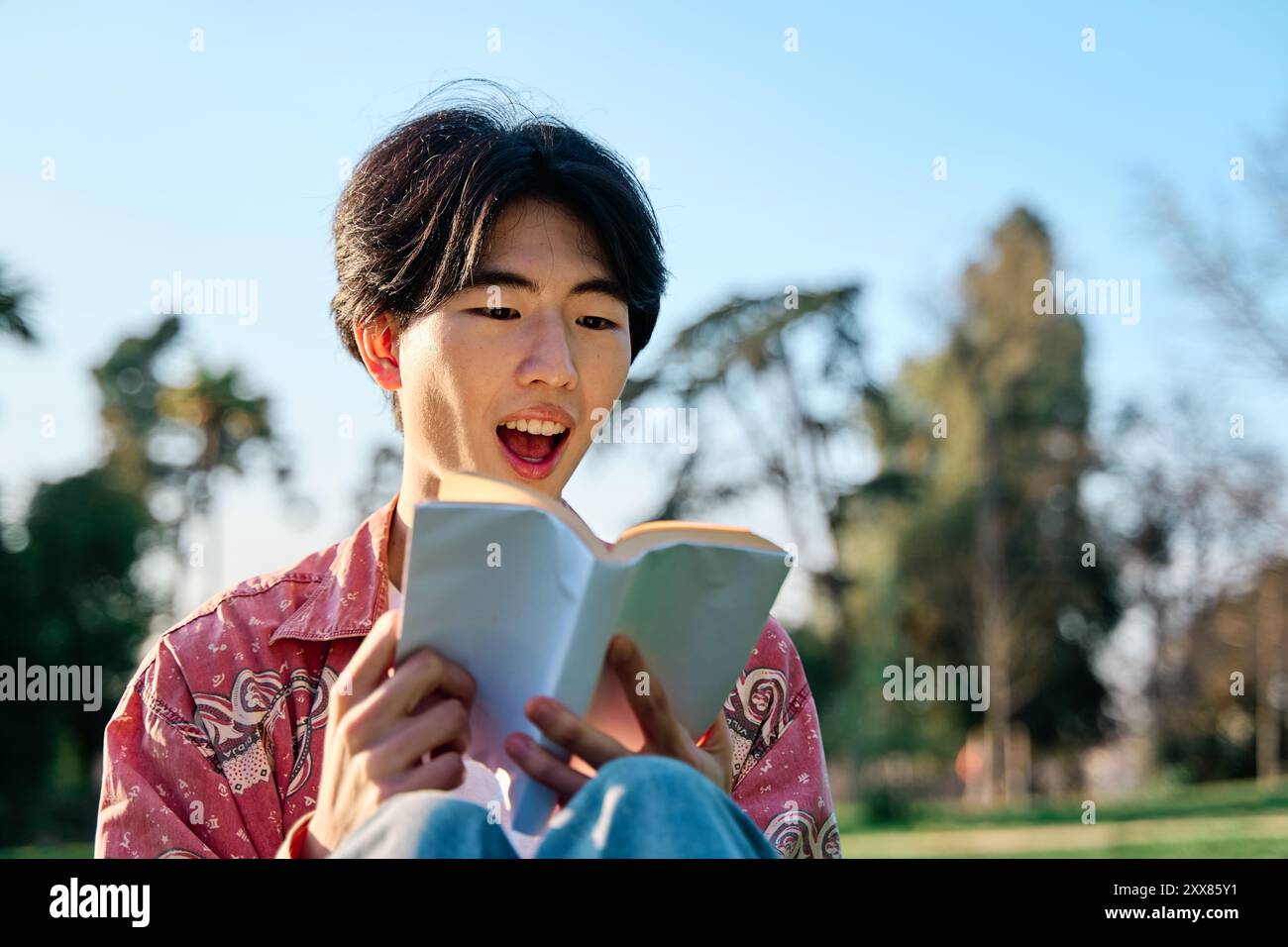 young asian man sitting and reading a book with a surprised look on his ...