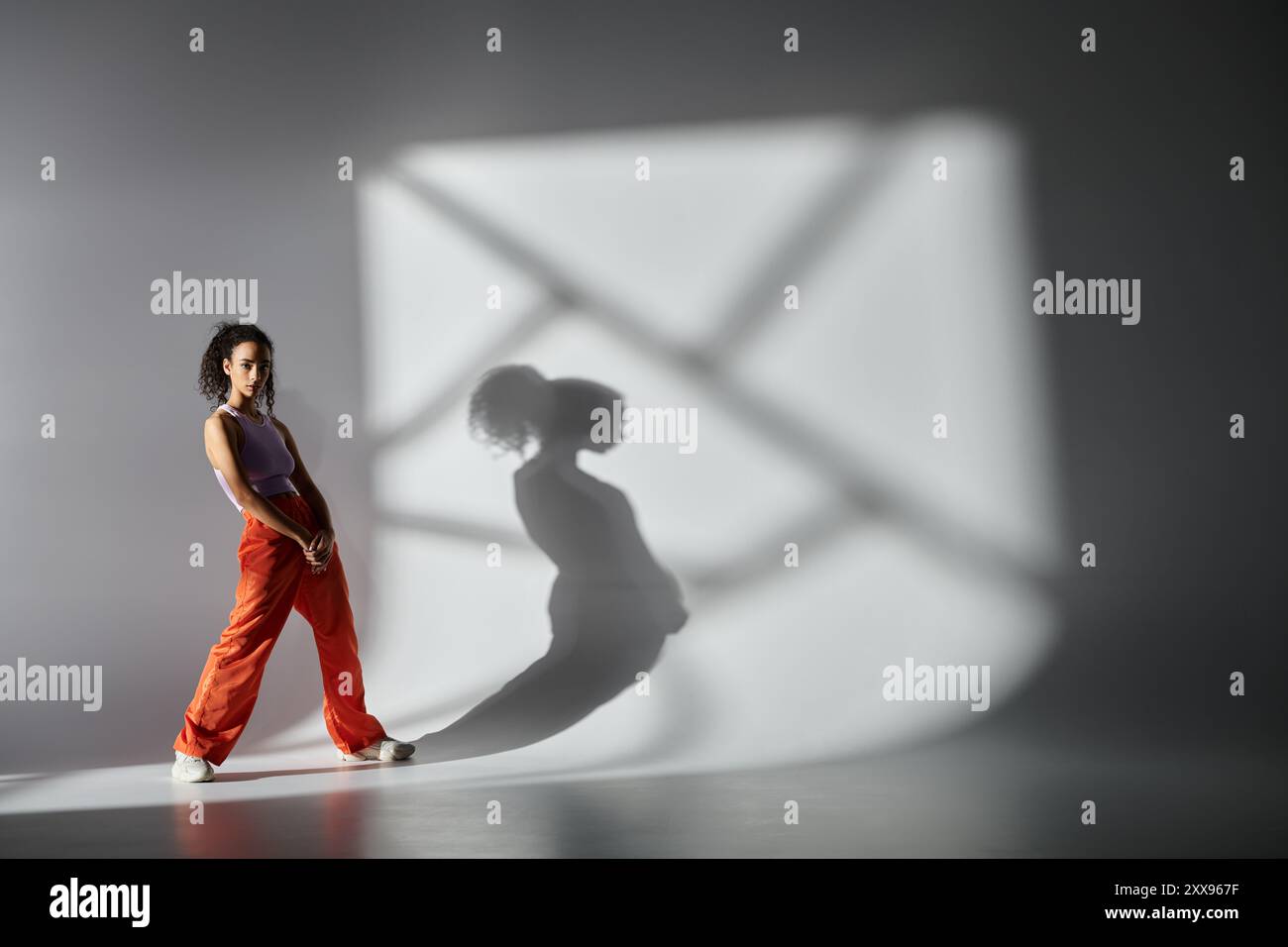 A young woman in trendy clothing poses in a studio with a grey backdrop ...