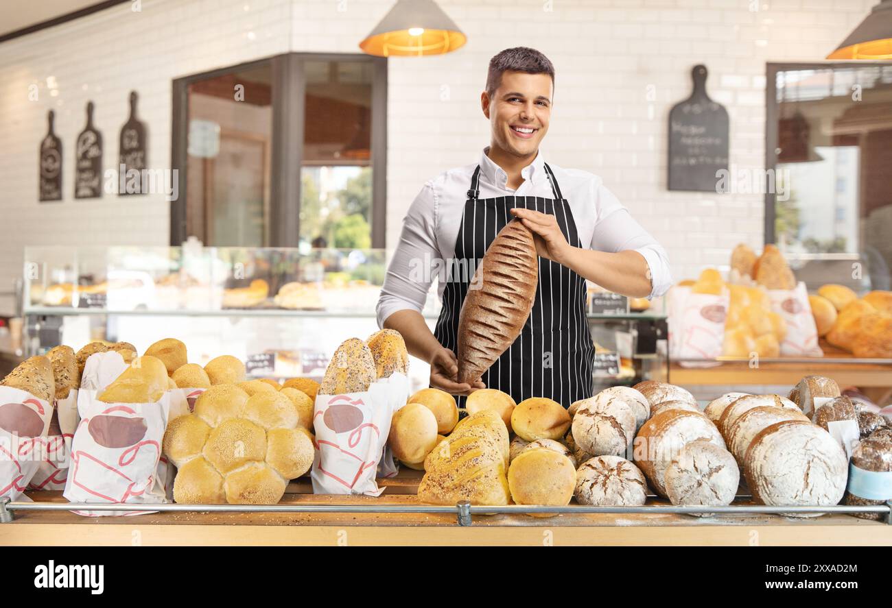 Young smiling male baker wearing an apron and holding a loaf of brown ...