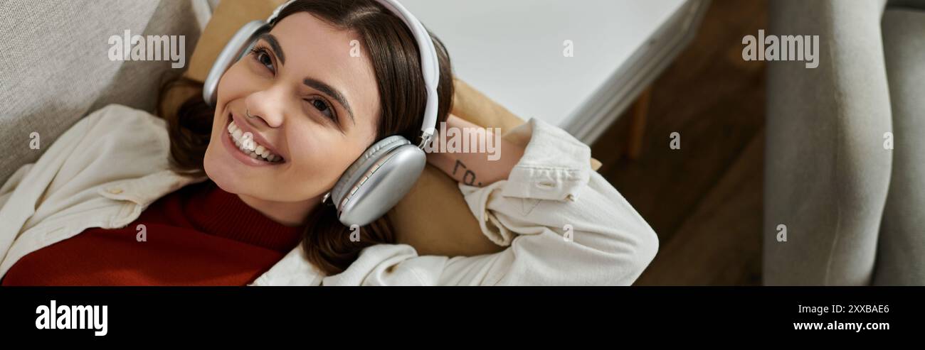 A young woman with headphones on, smiles while resting on her couch ...