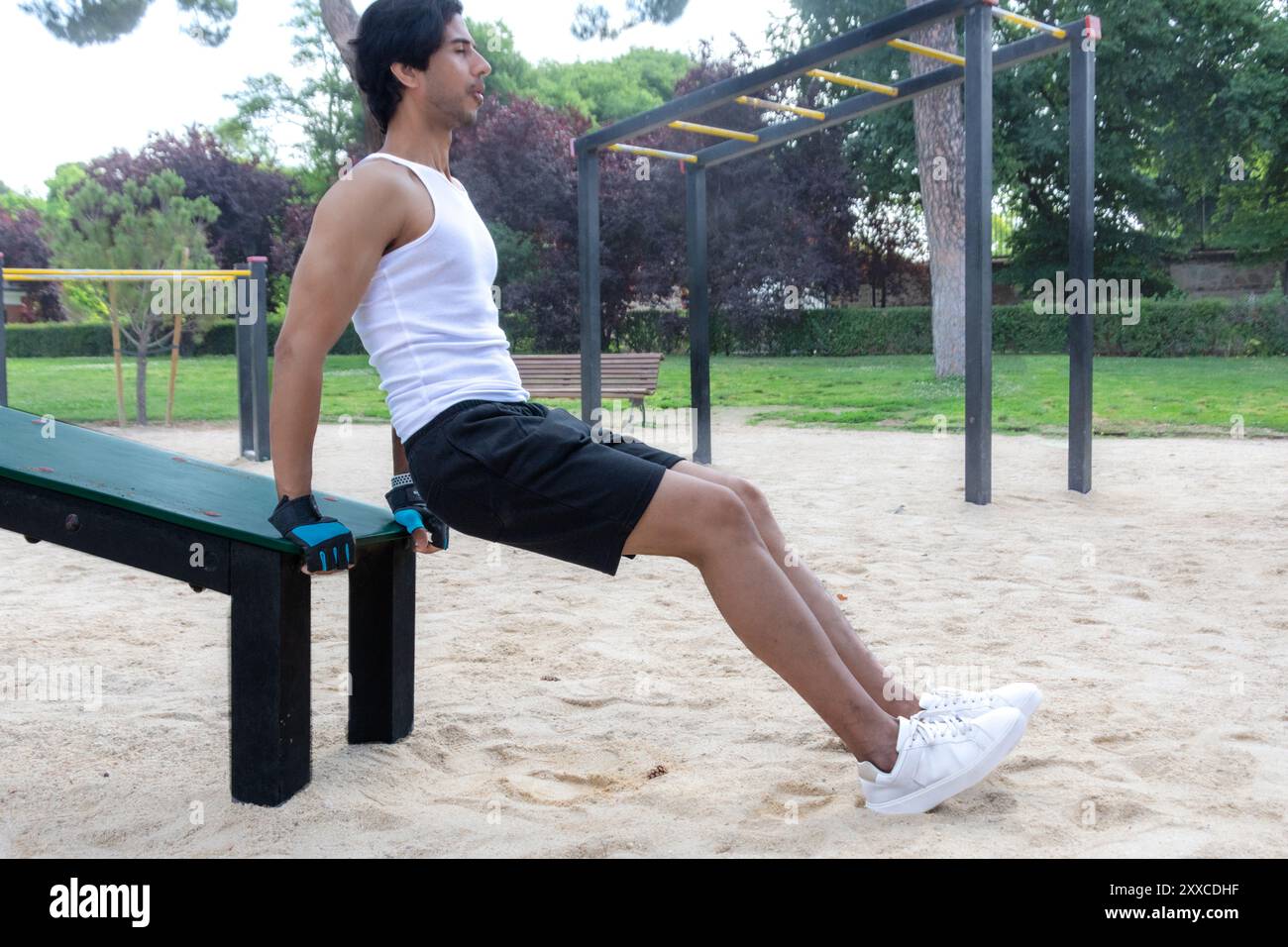 A man in a white tank top sits on a bench in a park. The bench is next ...