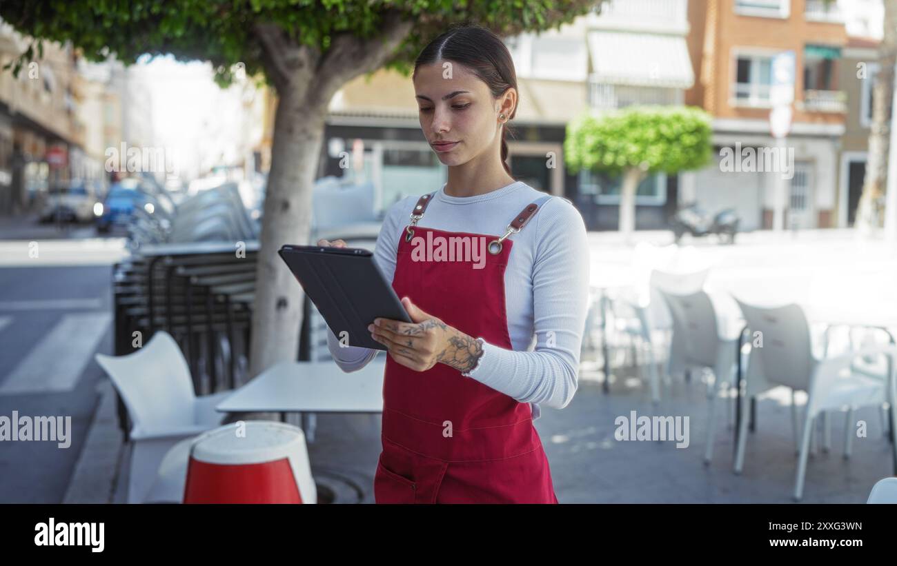 Young woman working at outdoor cafe terrace wearing red apron and using ...