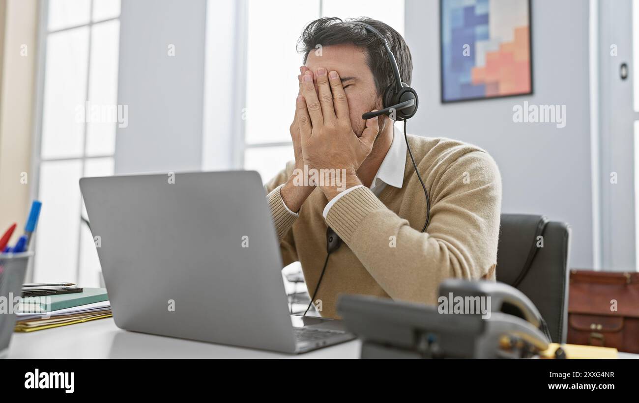 A stressed hispanic man with a beard wearing a headset covers his face ...
