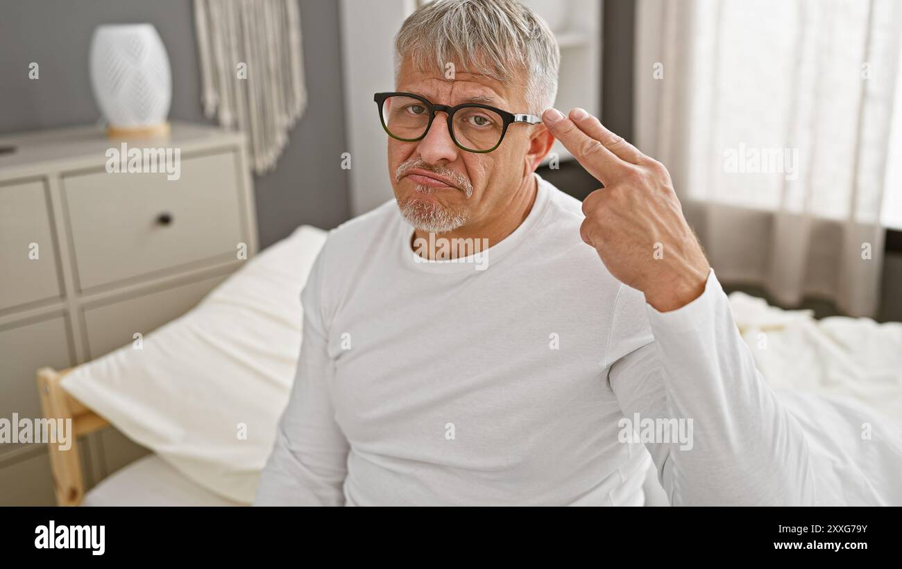 A perplexed, mature man in glasses sitting on a bed indoors, gesturing ...