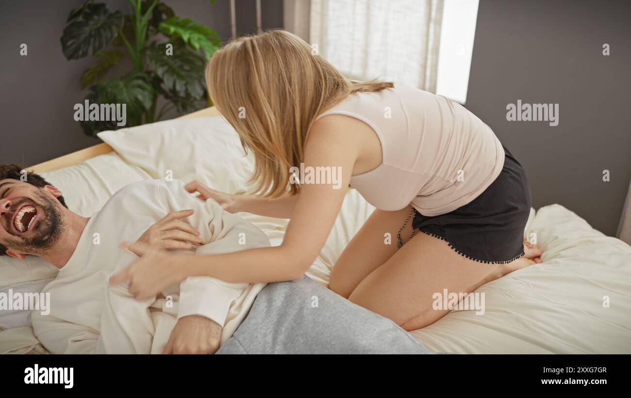Laughing man being tickled by playful woman on a bed in a cozy bedroom setting, reflecting joy ...