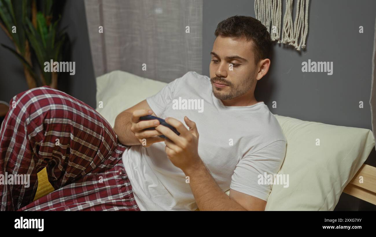 A handsome, young, hispanic man with a beard relaxes in a bedroom, lying on a bed and using his ...