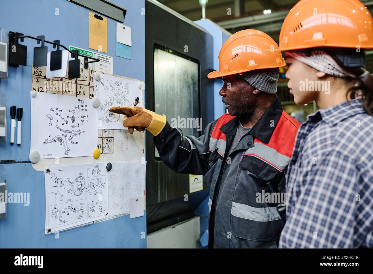 Two factory workers examining detailed blueprints pinned on a corkboard ...