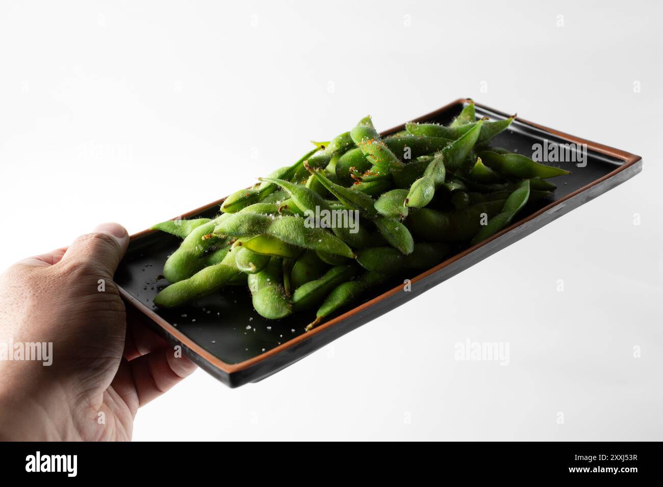 A view of a hand holding a plate of edamame, against of white ...